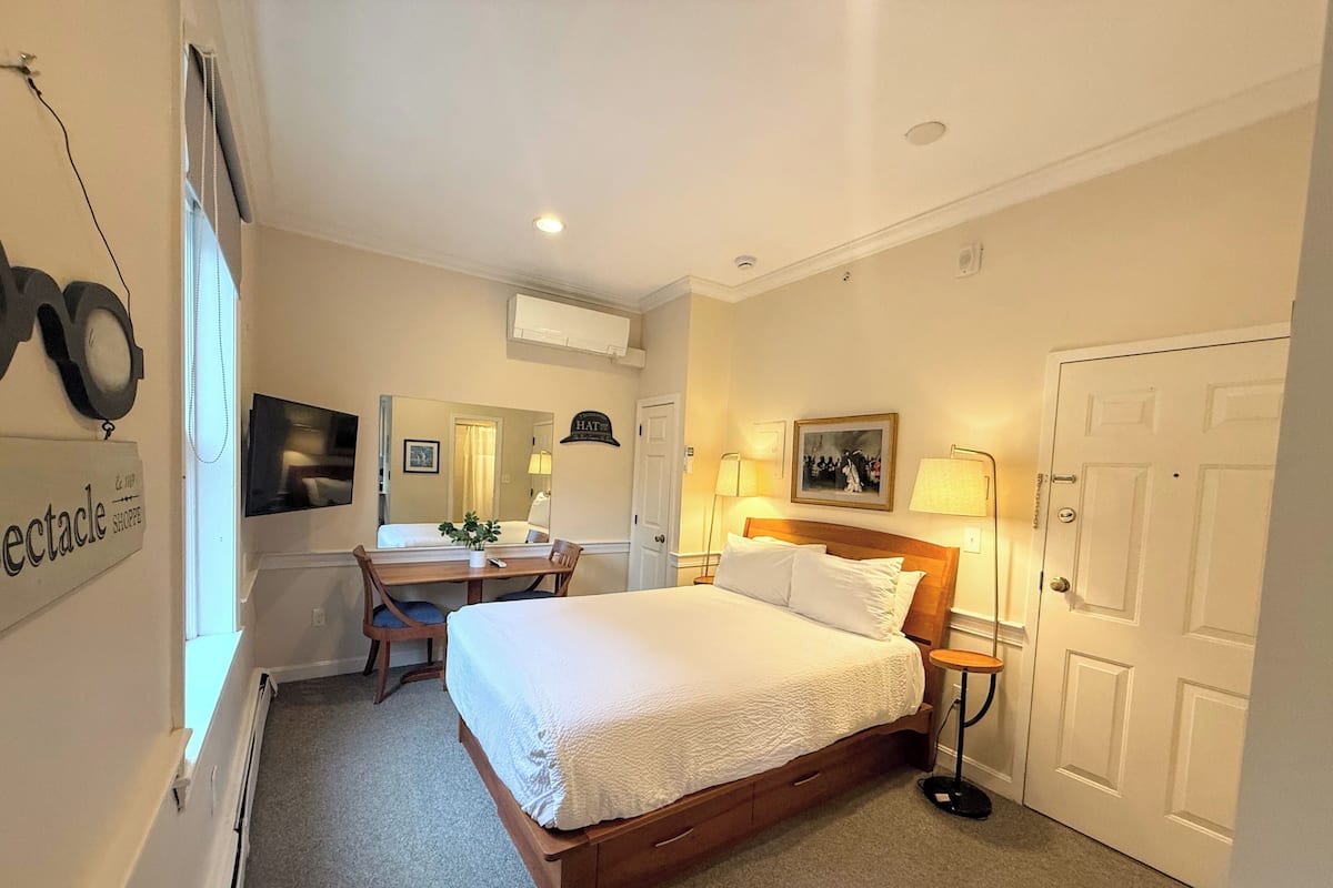 Quarto de hotel com cama, mesas de cabeceira, escrivaninha, cadeira, espelho e porta.