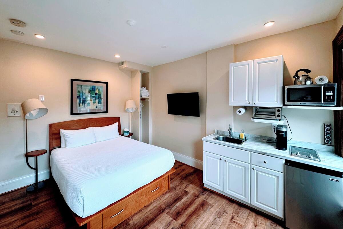 Quarto de hotel com cama, luminárias de cabeceira, TV, micro-ondas e uma pequena cozinha.