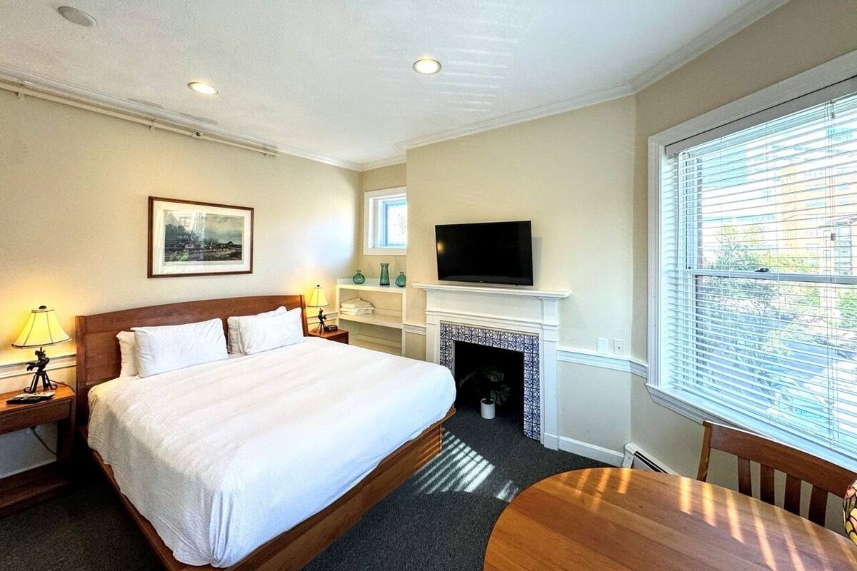 Quarto de hotel com cama grande, televisão sobre lareira, mesa pequena e janela com persianas.