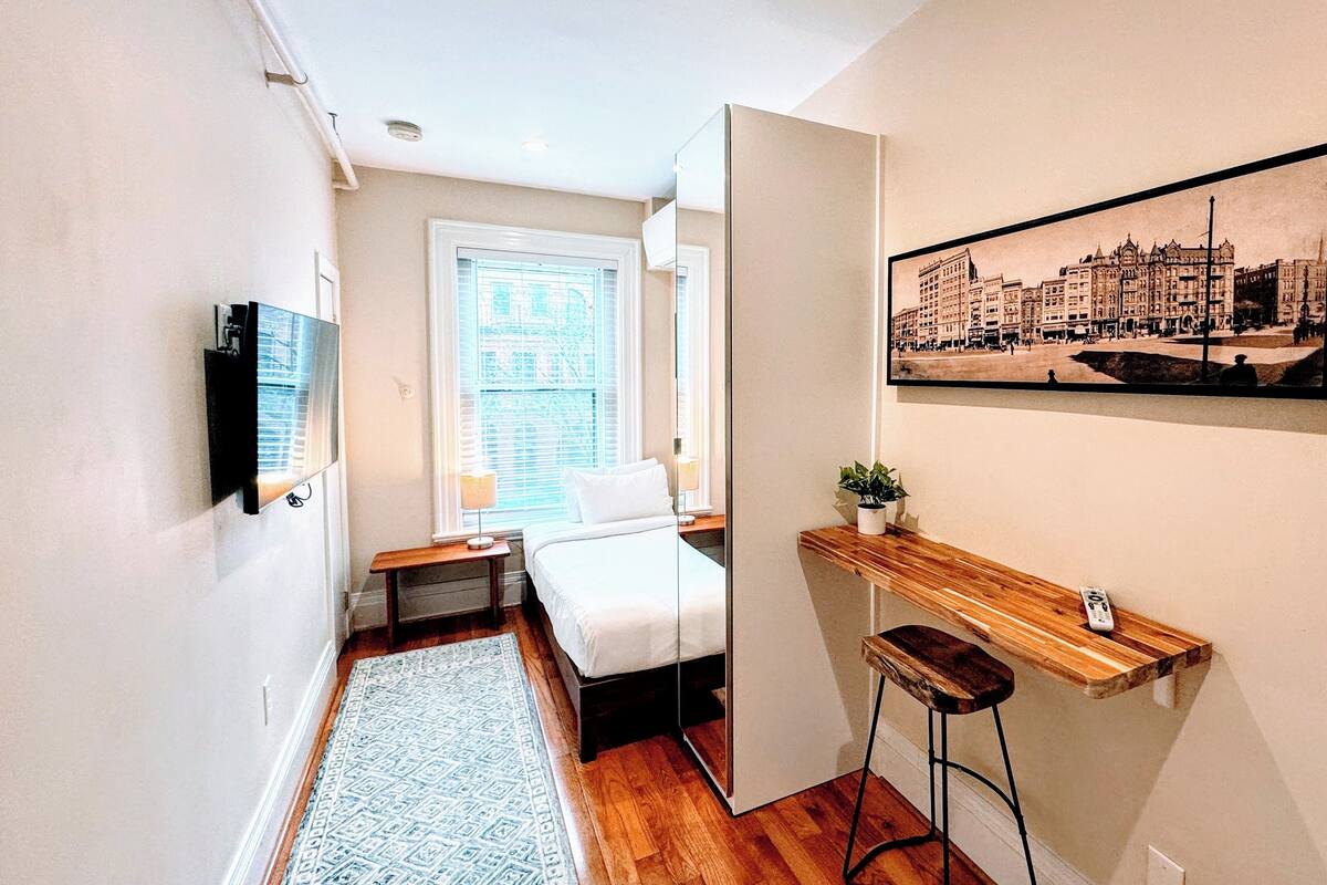 Quarto compacto com cama, mesa de cabeceira e um pequeno banquinho. Uma janela com persianas permite entrada de luz natural, e um quadro com vista urbana decora a parede.