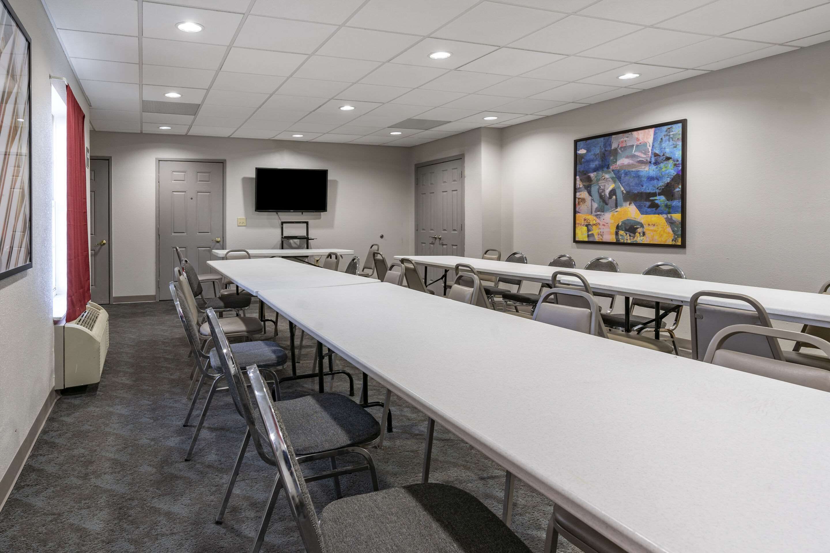 Uma sala de conferências com mesas compridas, cadeiras, uma TV de tela plana e um quadro na parede.