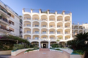 Hotel de vários andares com entrada central, varandas e vegetação ao redor.