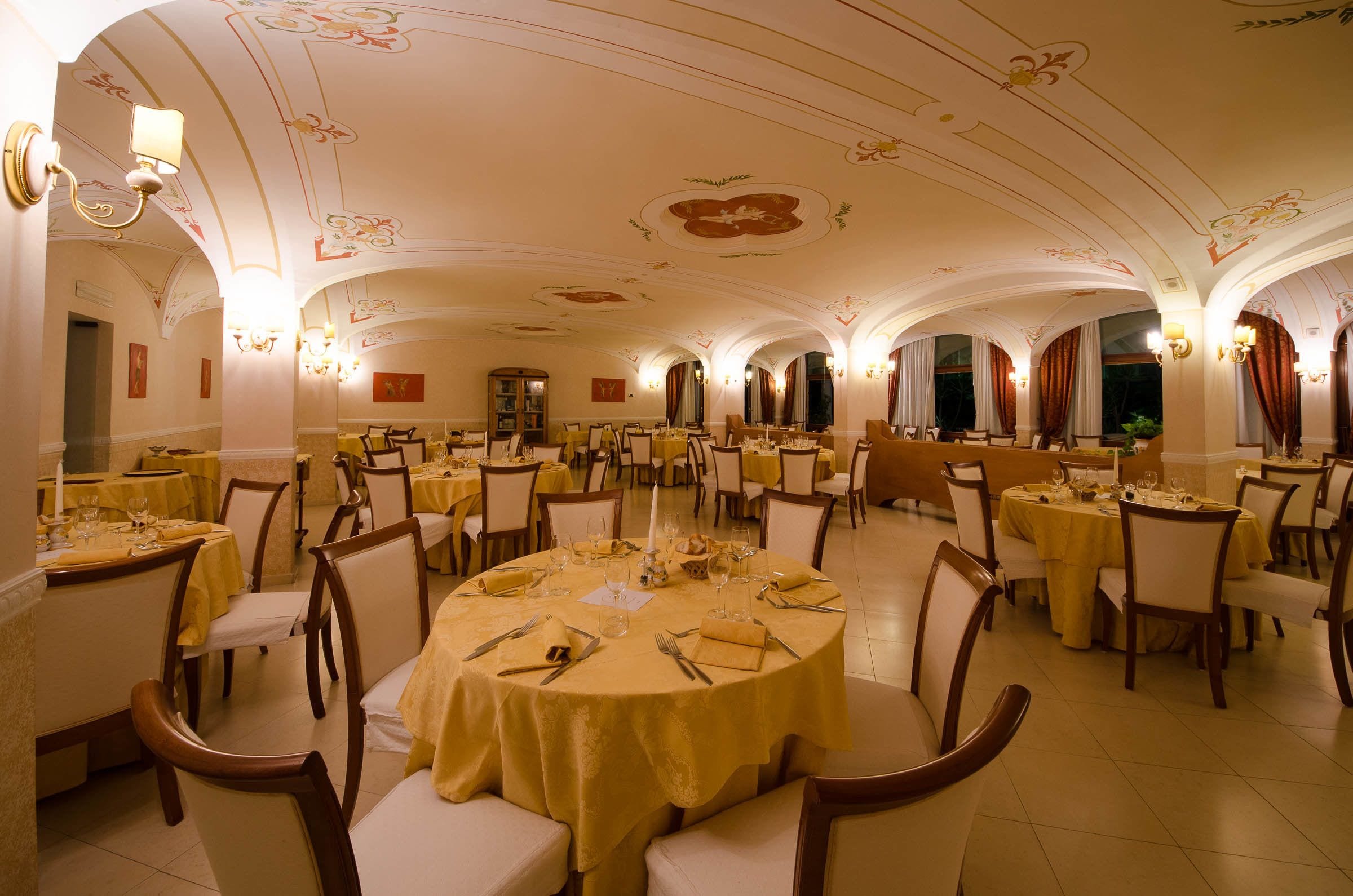 Um amplo salão de banquetes com várias mesas redondas dispostas para refeições, cada uma com cadeiras e louças.