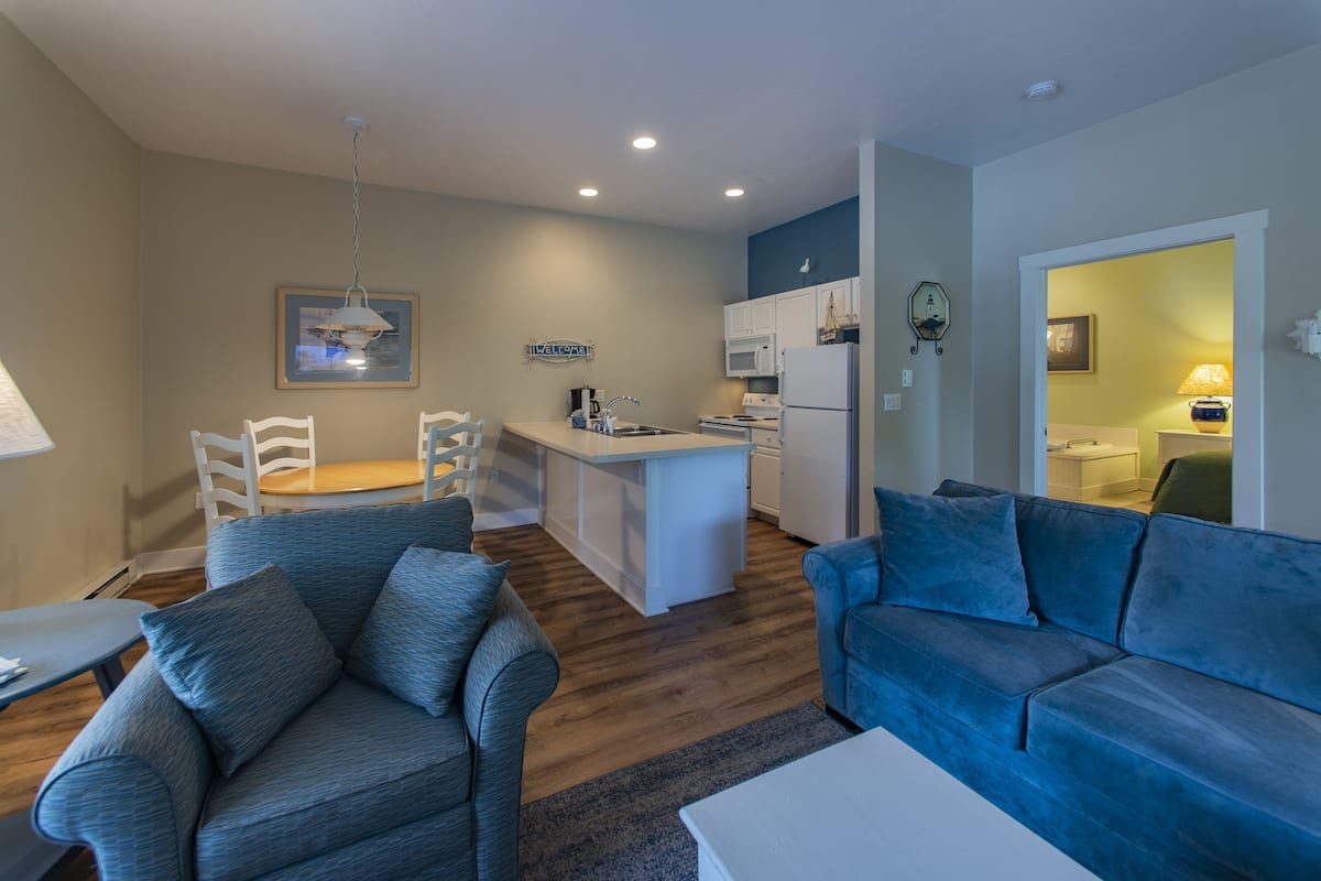 Uma sala de estar moderna com um sofá azul, um espaço de refeições com uma mesa branca e uma cozinha com armários e eletrodomésticos brancos.
