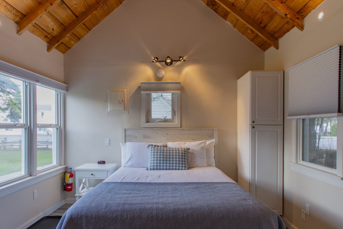 Um quarto com teto de madeira inclinado, uma cama com colcha xadrez, uma mesa de cabeceira, um pequeno armário, uma janela com persianas e uma porta.