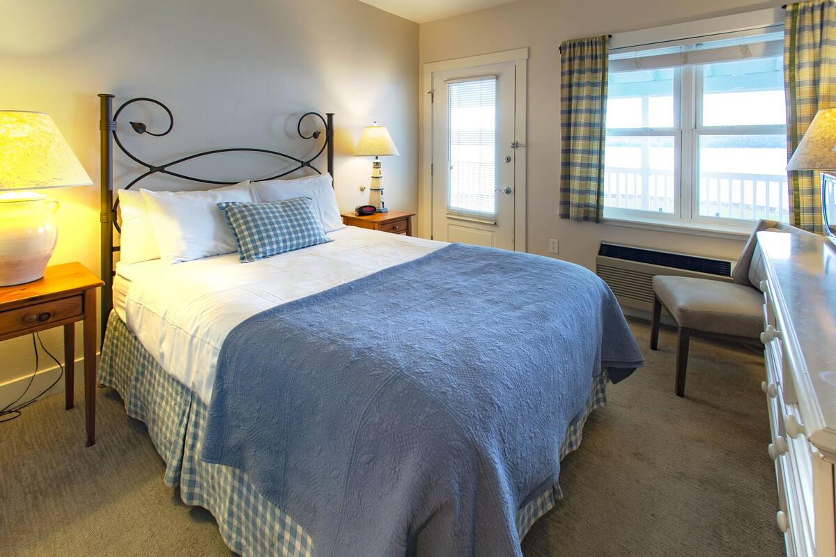 Cama bem arrumada com manta azul, duas luminárias de cabeceira, mesa de cabeceira de madeira e uma janela com cortinas.