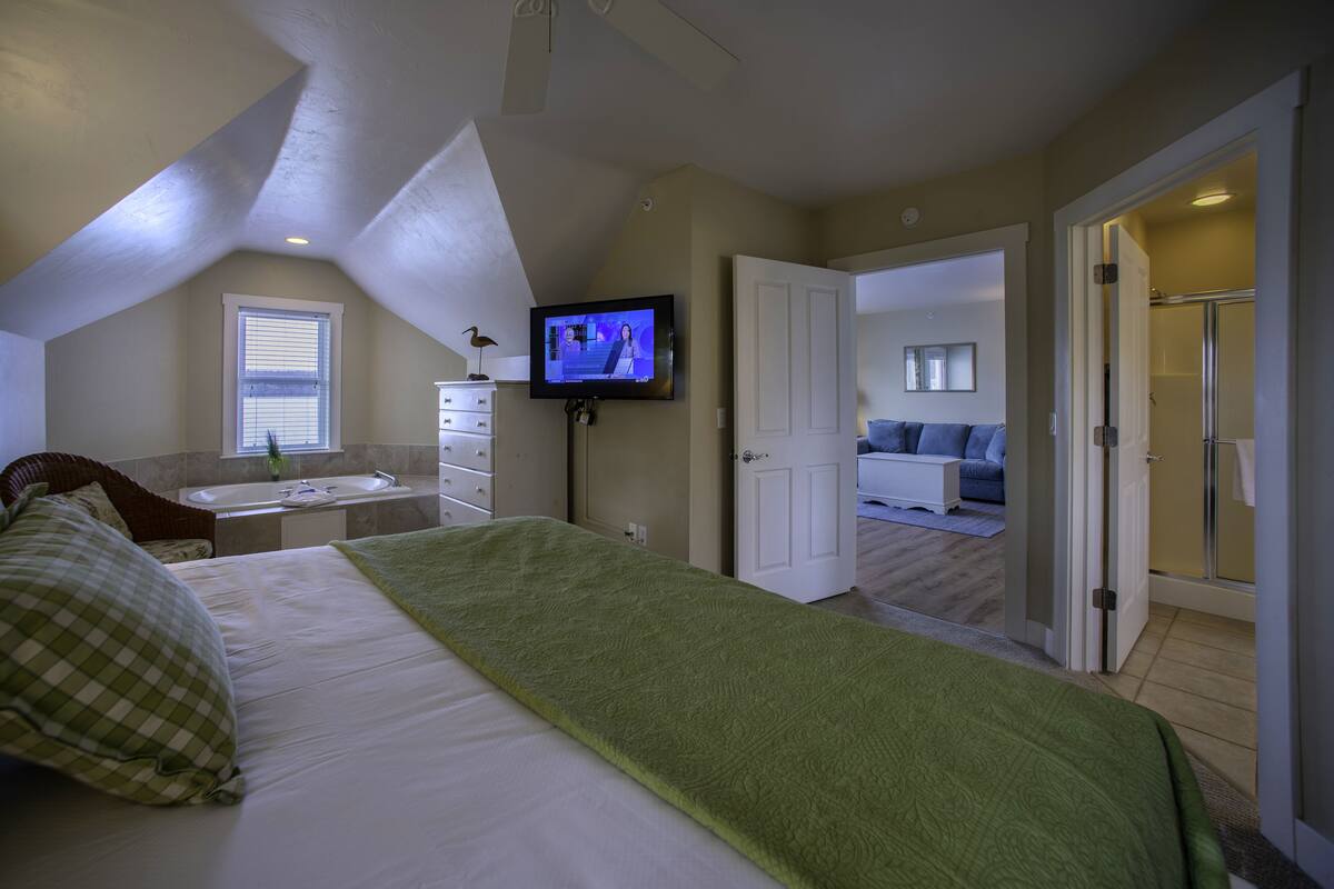 Um quarto com uma cama, uma televisão fixada na parede, um banheiro com pia e espelho, e uma porta aberta dando para outro cômodo.
