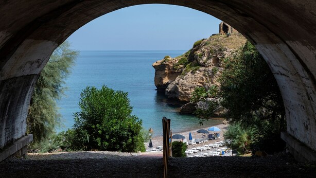 Vista através de um arco de pedra sobre uma área costeira com falésias rochosas, praia e guarda-sóis.