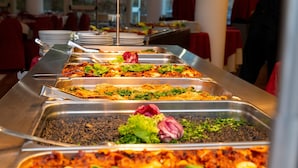 Buffet com diversos pratos, incluindo massas, saladas e opções com carne, dispostos em bandejas de metal.