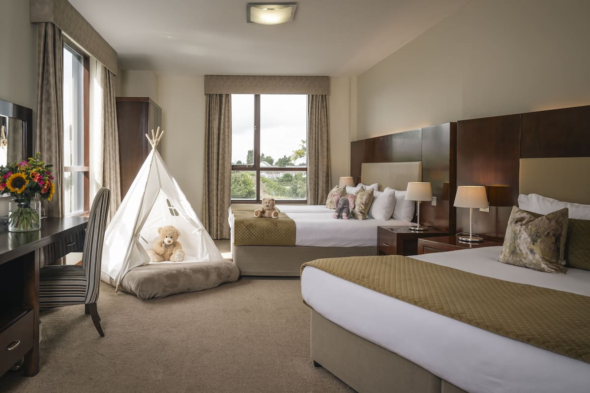 Quarto de hotel com duas camas, uma escrivaninha, uma cadeira, uma janela com cortinas, um ursinho de pelúcia e um tipi.