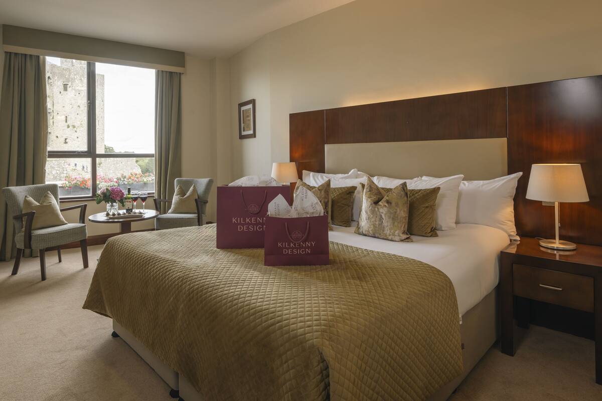 Quarto de hotel com uma cama grande, duas poltronas, uma pequena mesa com um vaso de flores e vista para um castelo pela janela.