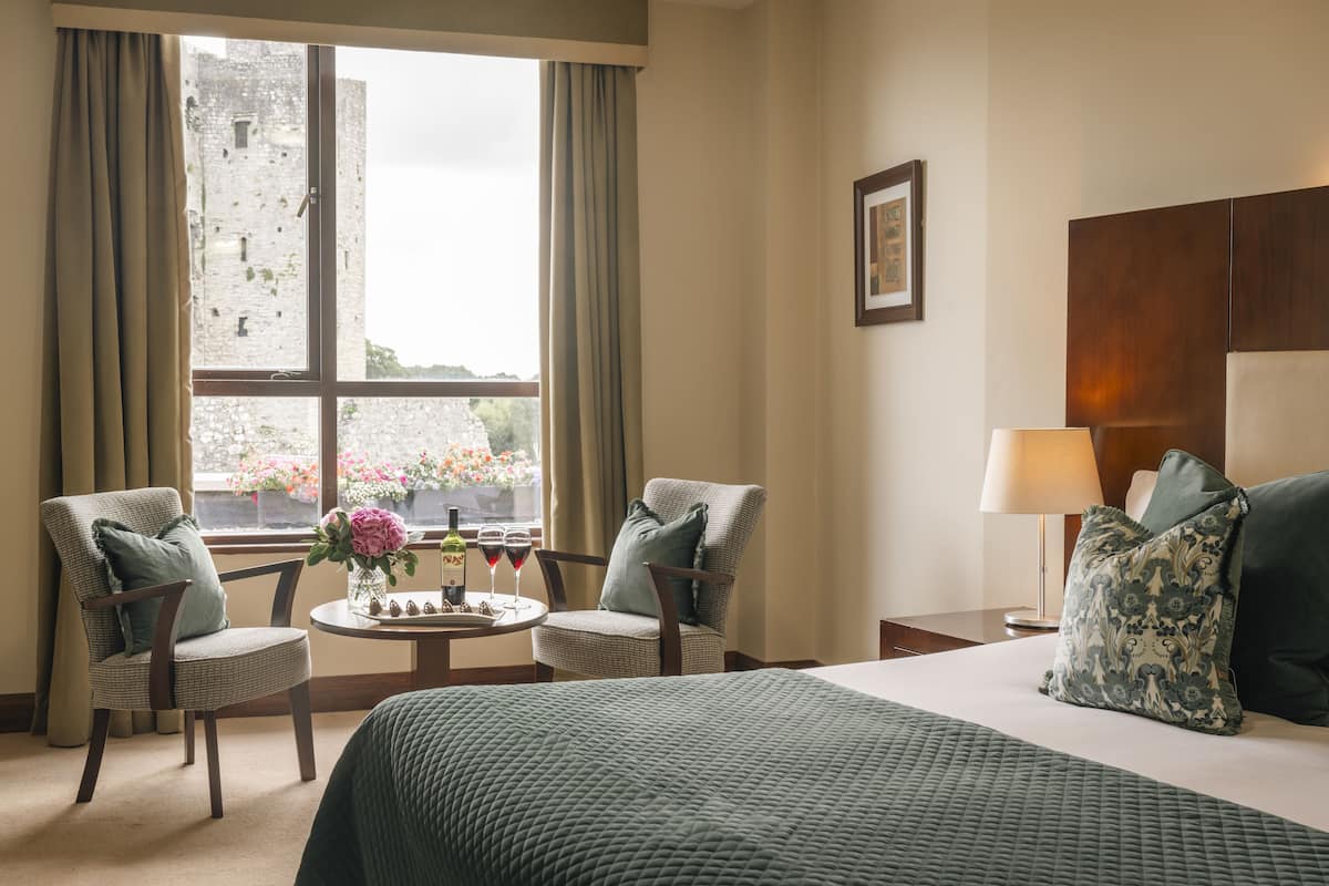 Quarto de hotel com cama grande, duas poltronas, uma mesinha com um vaso de flores e duas taças de vinho, e vista para um castelo pela janela.