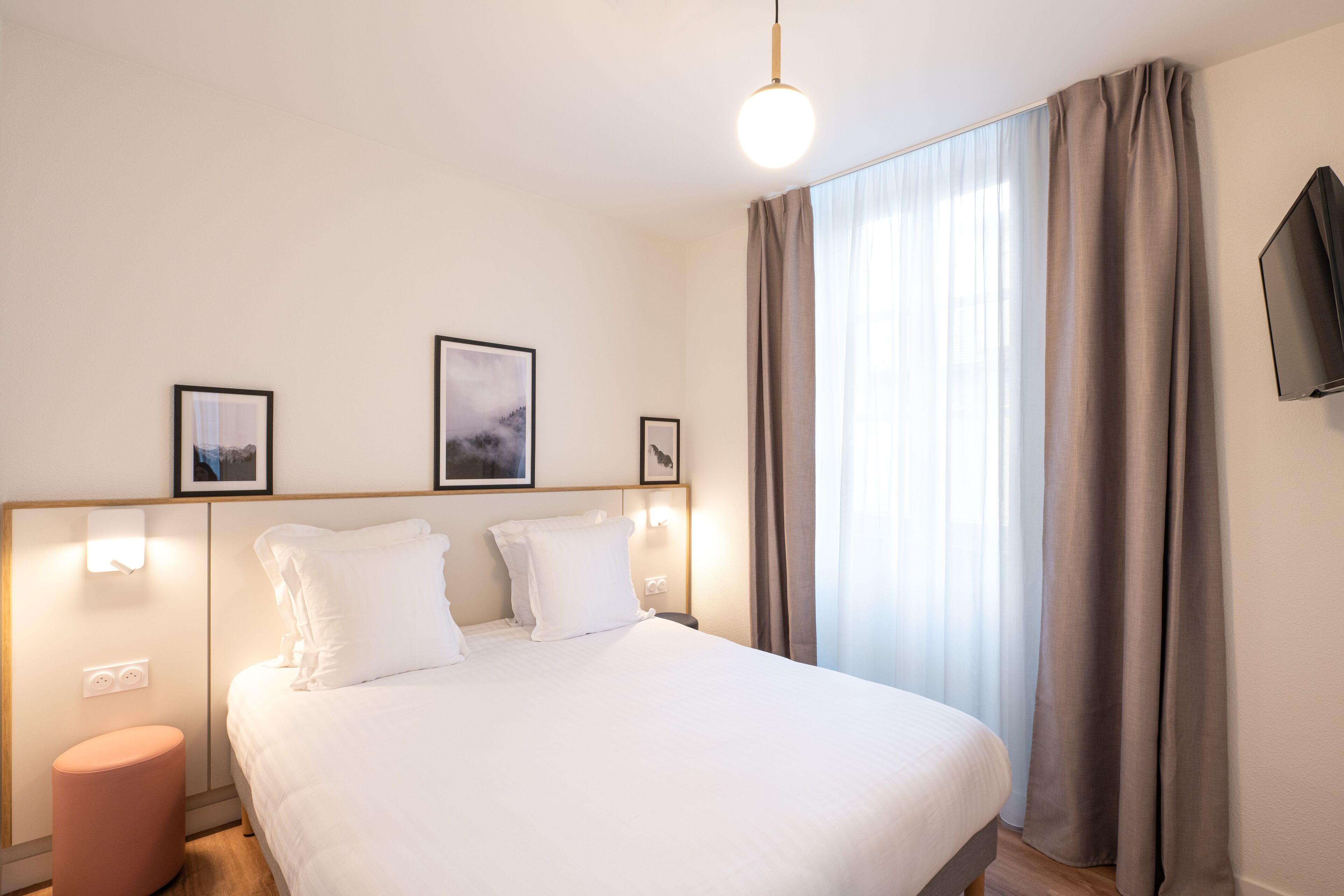 Quarto de hotel com uma cama grande, duas luminárias na parede, uma televisão de tela plana e uma janela com cortinas.