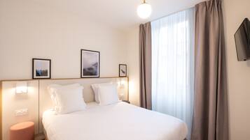 Quarto de hotel com uma cama grande, duas luminárias na parede, uma televisão de tela plana e uma janela com cortinas.