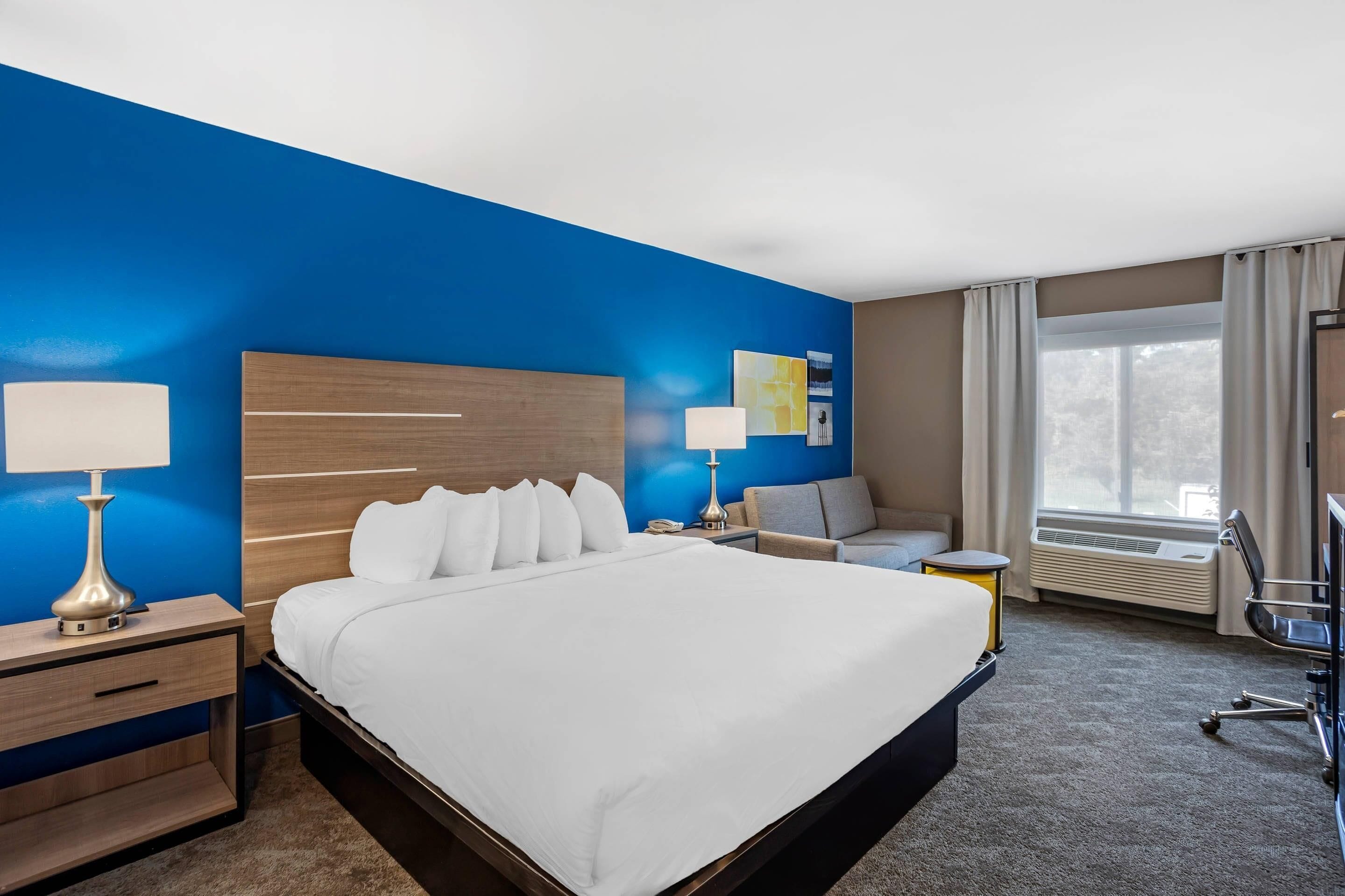 Quarto de hotel com uma cama grande, uma escrivaninha com candeeiro, uma cadeira, uma mesinha e uma janela com cortinas.