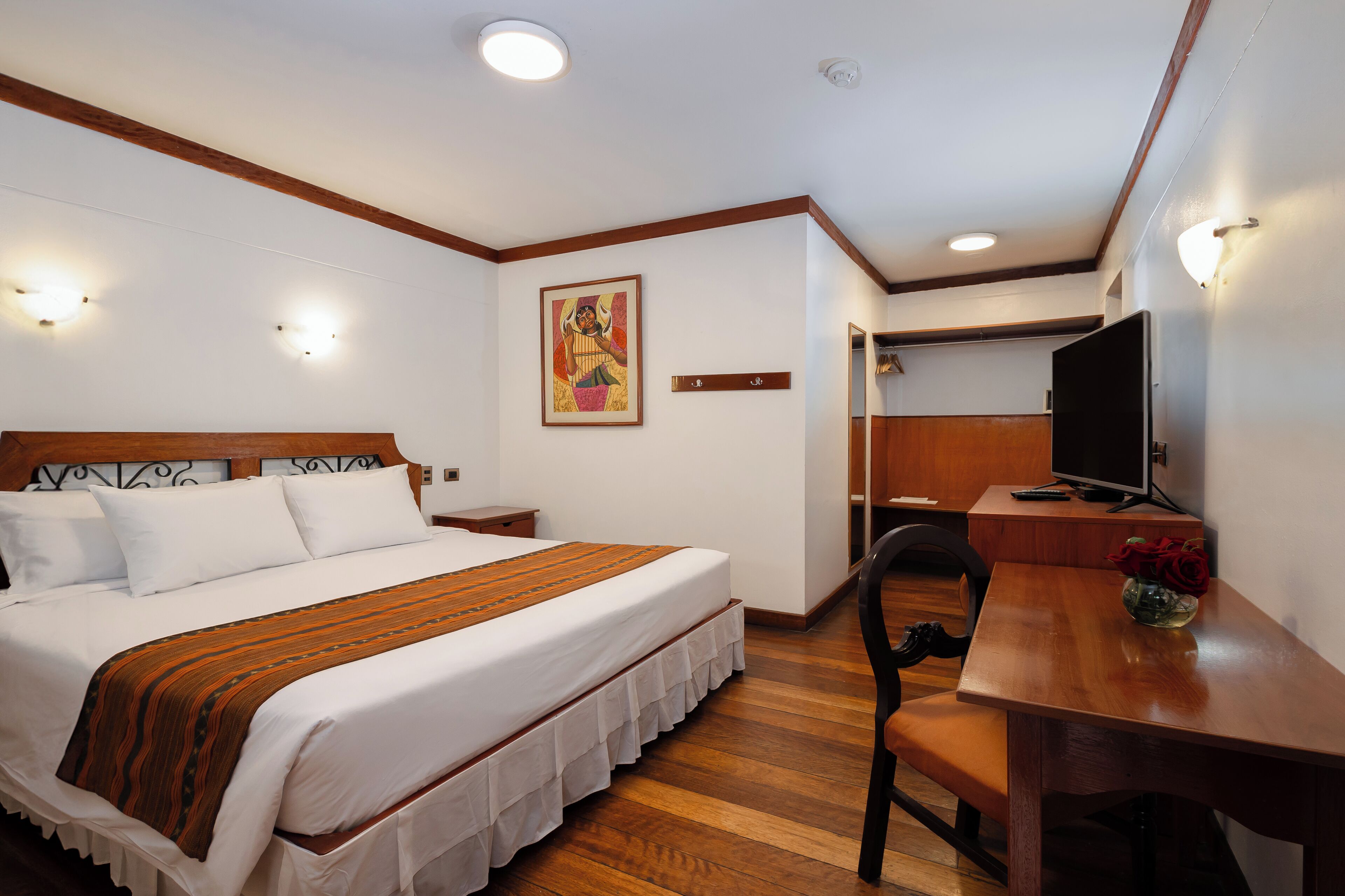 Quarto de hotel com uma cama grande, uma escrivaninha com cadeira, televisão e um quadro na parede.