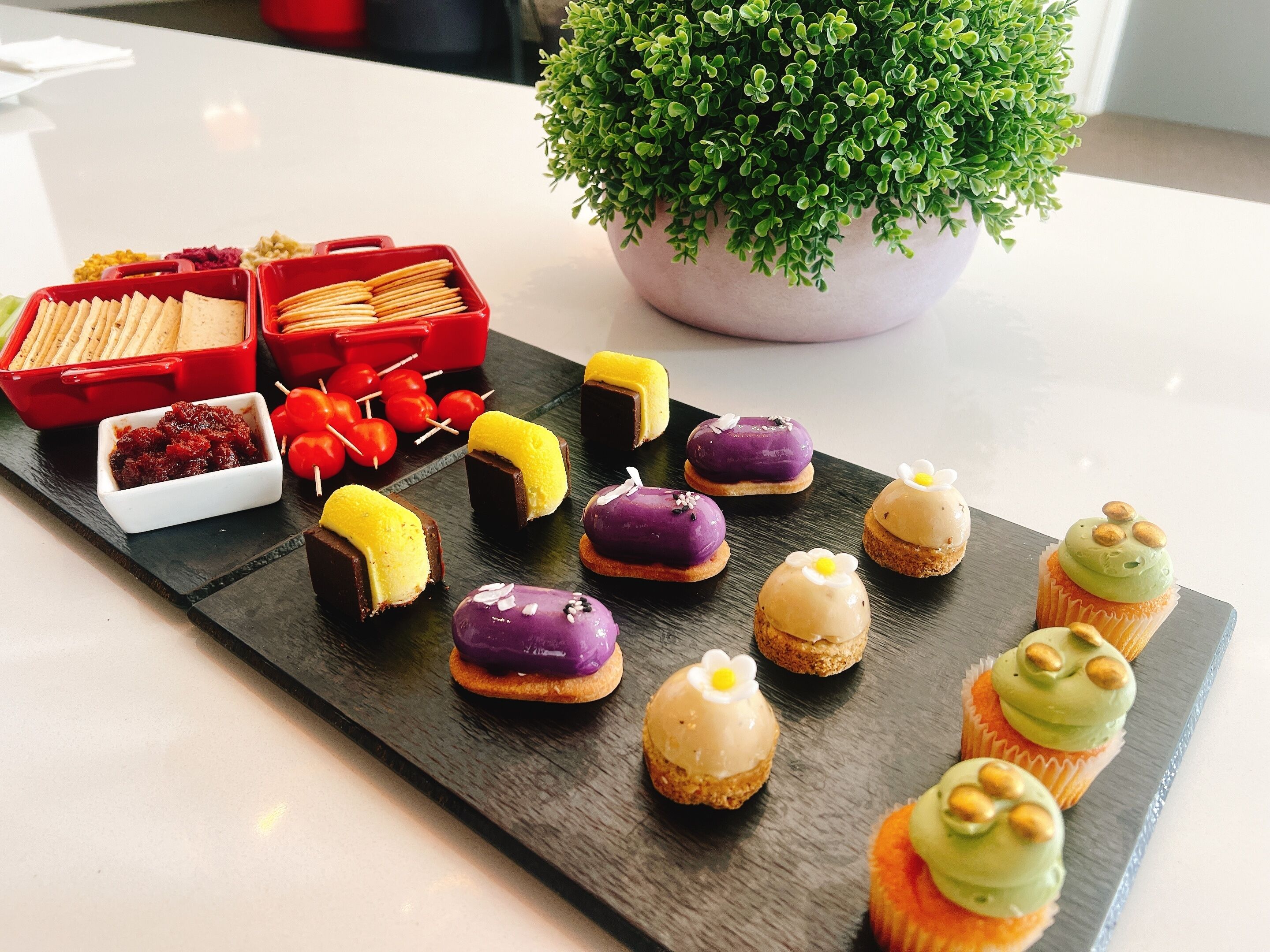 Diversos doces em um suporte de ardósia, incluindo cupcakes e guloseimas com cobertura de chocolate, com uma planta em vaso ao fundo.