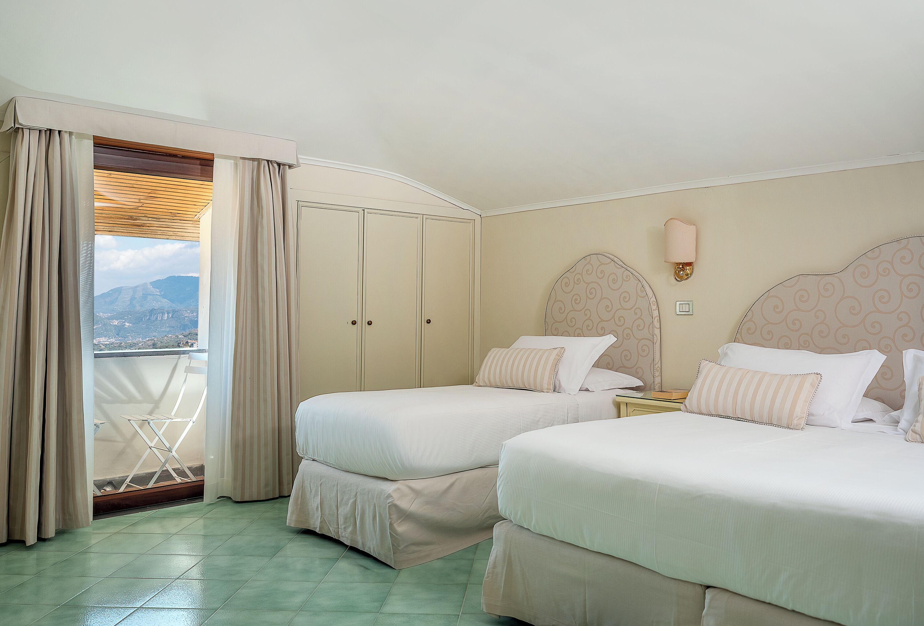 Quarto de hotel com duas camas, varanda com cortinas, uma mesa pequena e uma luminária na parede.