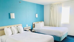 Quarto de hotel com duas camas, parede azul, janela com cortinas e mesa de cabeceira com telefone e abajur.
