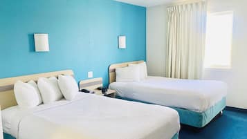Quarto de hotel com duas camas, parede azul, janela com cortinas e uma pequena mesa com telefone e abajur.