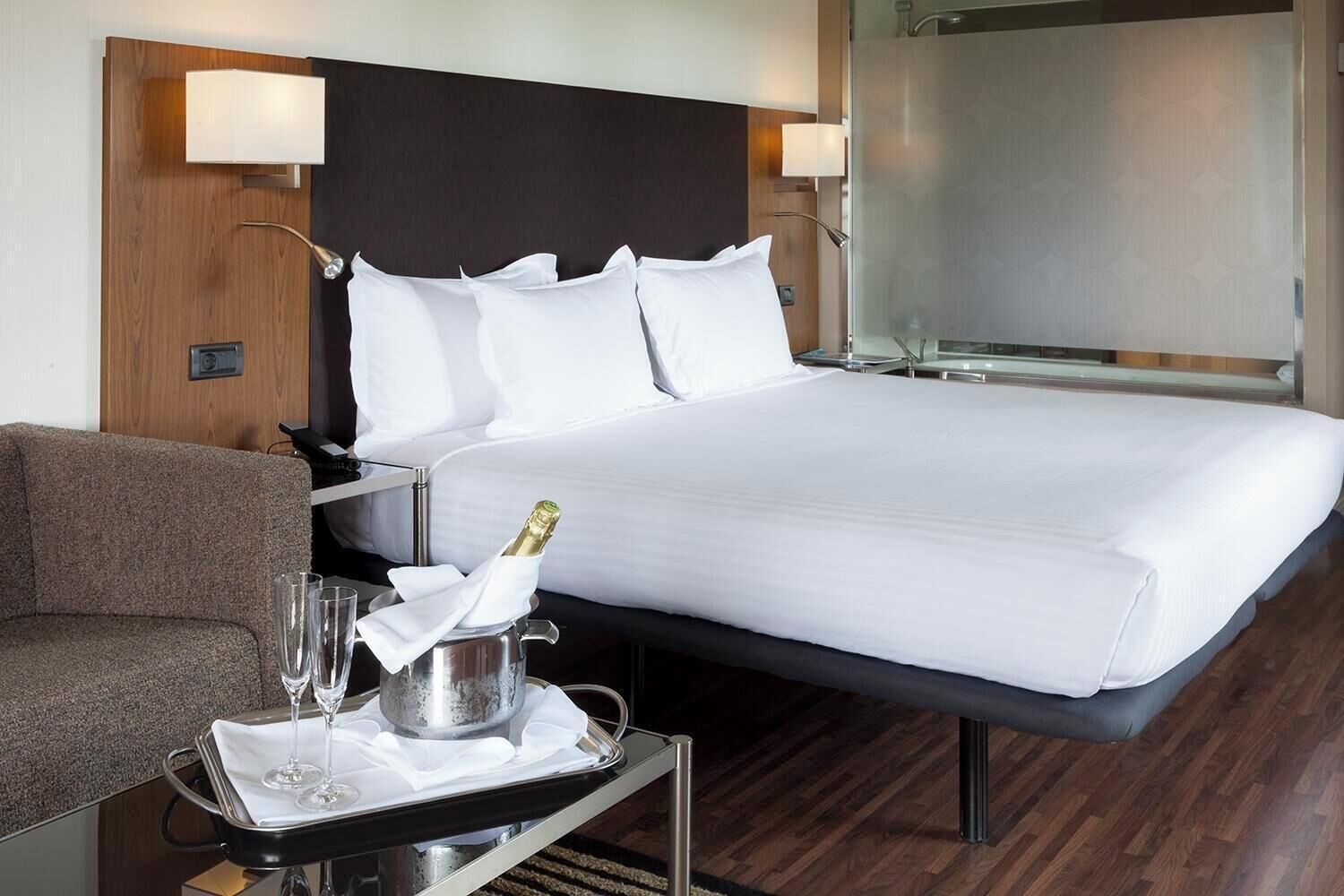 Quarto de hotel com uma cama grande, mesa de cabeceira com uma garrafa de champanhe, copos e um tabuleiro, cabeceira de madeira e área de chuveiro.