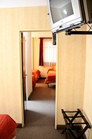 Quarto de hotel com televisão na parede, cama com roupa de cama laranja e um banquinho de madeira.