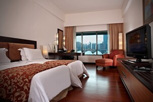 Quarto de hotel com uma cama grande, uma escrivaninha com cadeira, televisão e vista para a paisagem urbana pelas grandes janelas.