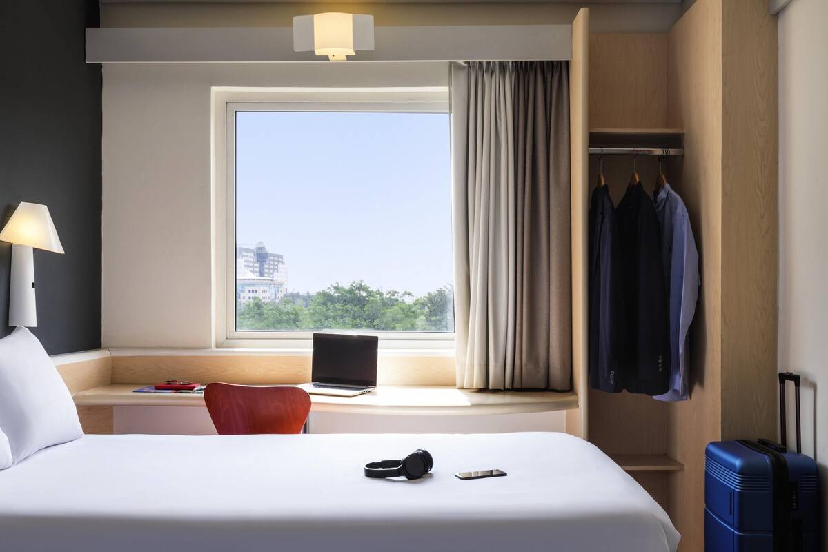 Quarto de hotel com uma cama, uma escrivaninha com um laptop, uma cadeira, uma mala e um guarda-roupa.