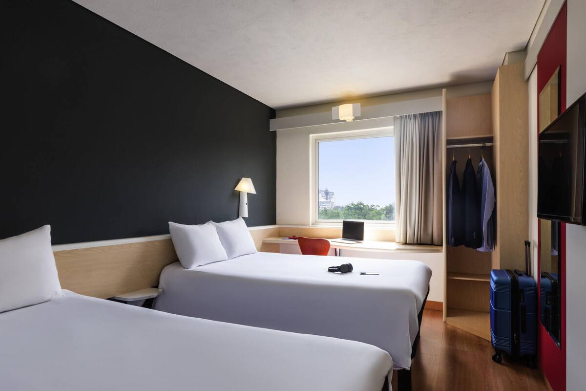 Quarto de hotel com duas camas, uma escrivaninha com um laptop, uma televisão e um guarda-roupa.