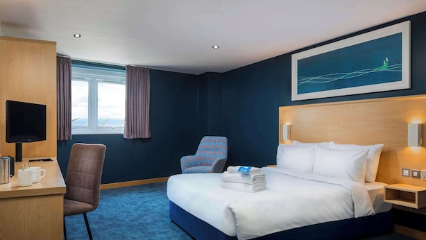 Quarto de hotel com cama, secretária com televisão, cadeira e janela com cortinas.