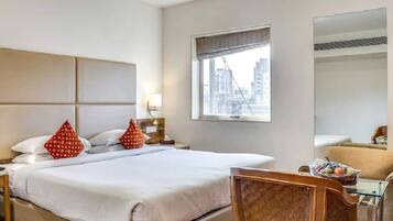Quarto de hotel com uma cama grande, uma cadeira, uma mesa pequena com uma cesta de frutas, um criado-mudo com abajur e uma janela com vista para a cidade.