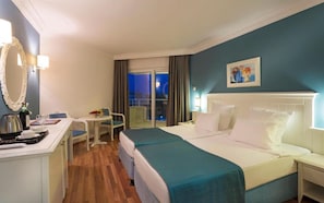 Quarto de hotel com duas camas, uma escrivaninha com cadeira, uma mesinha com uma tigela de frutas, um espelho e um quadro na parede.