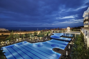 Uma grande piscina iluminada com várias pistas de natação, rodeada por espreguiçadeiras e guarda-sóis, com o céu noturno limpo e uma vista distante do oceano.