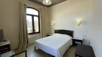Quarto de hotel com cama, mesas de cabeceira, escrivaninha com cadeira, janela com cortinas e luminária no teto.