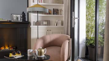 Um quarto aconchegante com lareira, uma poltrona confortável, uma mesinha com um bule e uma moldura, e uma estante com livros e objetos decorativos.