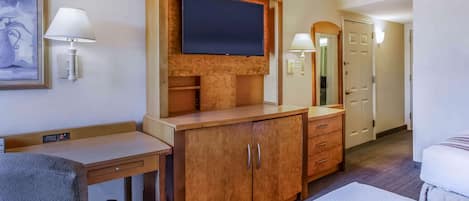 Quarto de hotel com escrivaninha de madeira, televisão fixada em painel de madeira, cama com roupa de cama listrada e porta para outro cômodo.