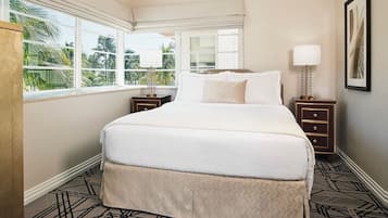 Cama bem arrumada com edredom branco e travesseiros, mesa de cabeceira com abajur e janela com vista para palmeiras.