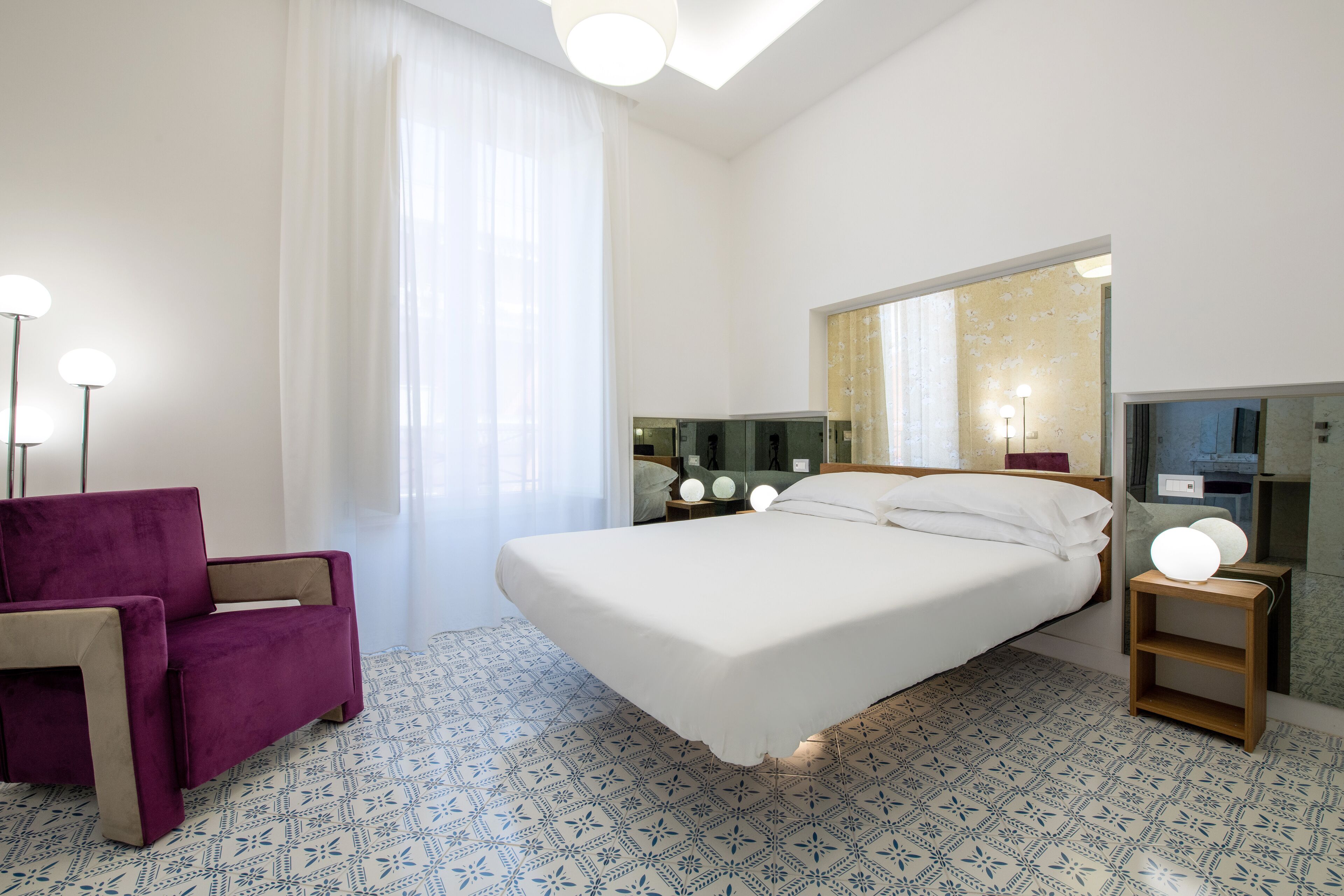 Quarto de hotel moderno com uma cama grande, uma poltrona roxa, uma mesinha de cabeceira com uma luminária e um espelho amplo.