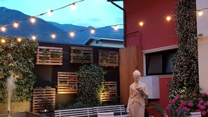 Uma área externa com mesas e bancos de madeira, rodeada por plantas em vasos e uma estátua.