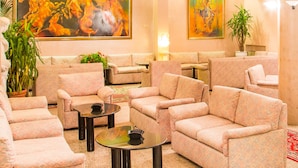 Um hall amplo com várias áreas de estar, incluindo sofás e poltronas, dispostas ao redor de uma mesa de centro. Há duas grandes pinturas nas paredes, e um vaso de plantas é visível no canto.