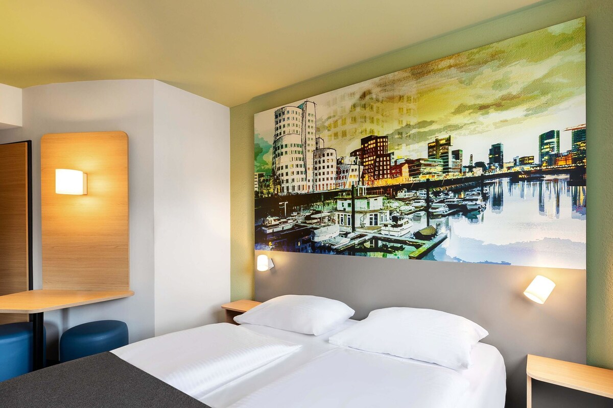 Quarto de hotel com uma cama, uma mesa e uma grande pintura de uma paisagem urbana na parede.