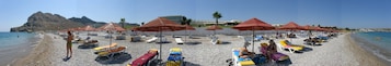 Uma praia com espreguiçadeiras e guarda-sóis, um litoral rochoso e um edifício ao fundo.