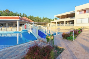 Piscina com uma ponte, rodeada por uma área ajardinada com plantas e um edifício com telhado em azulejos.