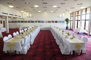 Uma sala de banquetes com longas mesas preparadas para um evento formal, cadeiras brancas e um tapete vermelho.