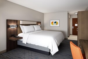 Quarto de hotel moderno com uma cama grande, mesa de cabeceira com telefone, prateleira pequena e uma obra de arte na parede.