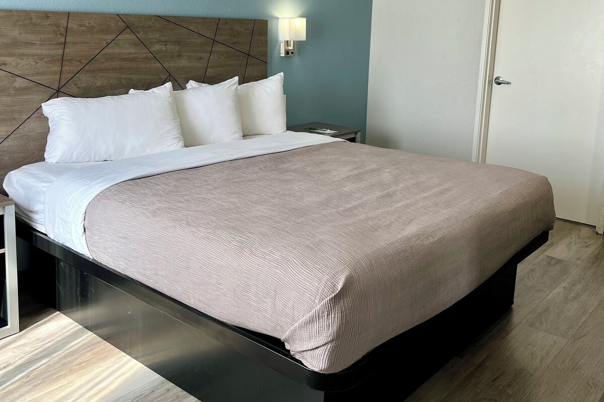Quarto de hotel moderno com cama grande, cabeceira de madeira, mesa de cabeceira, luminária na parede e uma porta.