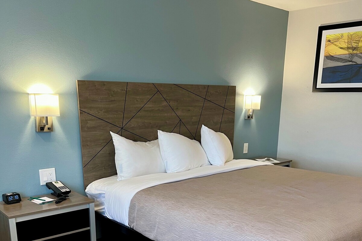 Quarto de hotel moderno com cabeceira de madeira, cama grande com roupa de cama branca, mesa de cabeceira com telefone e relógio, e luminárias fixadas na parede.