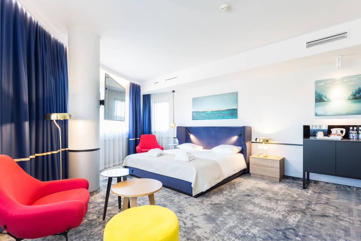 Quarto de hotel moderno com uma cama grande, uma poltrona vermelha, um banquinho amarelo, uma mesa redonda pequena e uma mesa de cabeceira com abajur.