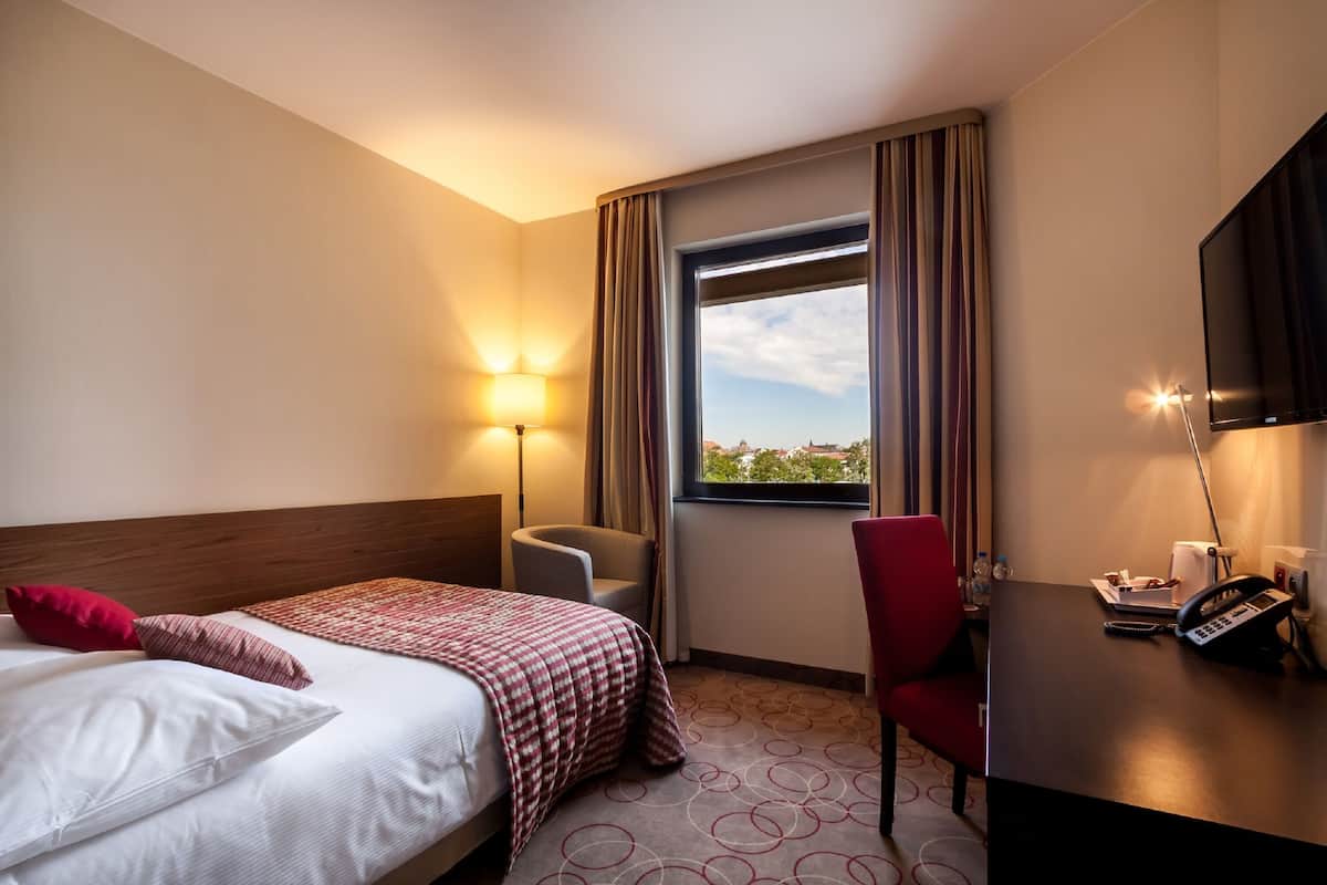 Quarto de hotel com cama, escrivaninha com telefone, cadeira, luminária e janela com cortinas.