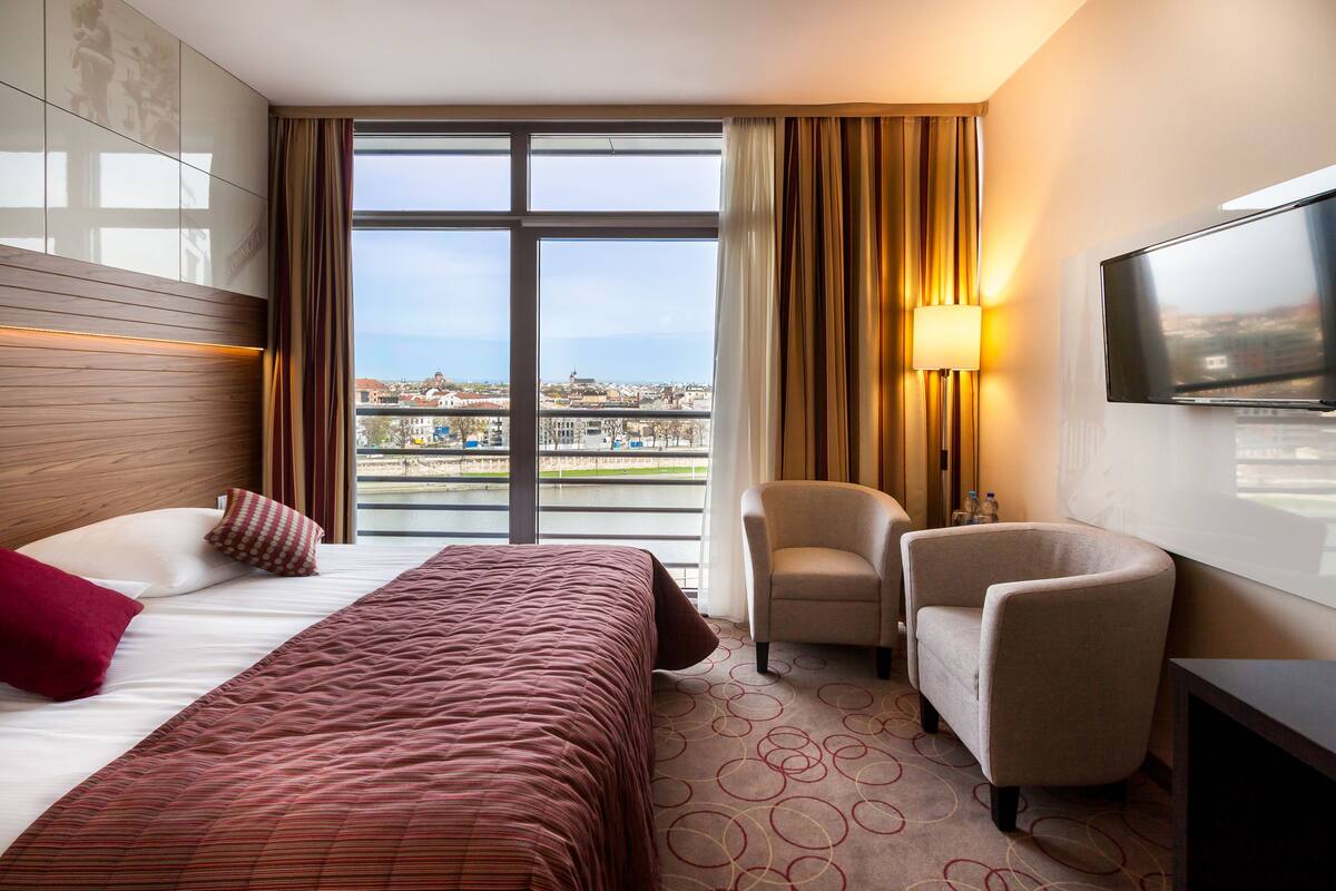 Quarto de hotel com uma cama grande, duas poltronas, uma televisão e vista para a paisagem urbana.